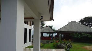 Uma área externa coberta com piso de azulejos, um pequeno jardim com pedras e plantas, e um edifício com telhado de azulejos ao fundo.