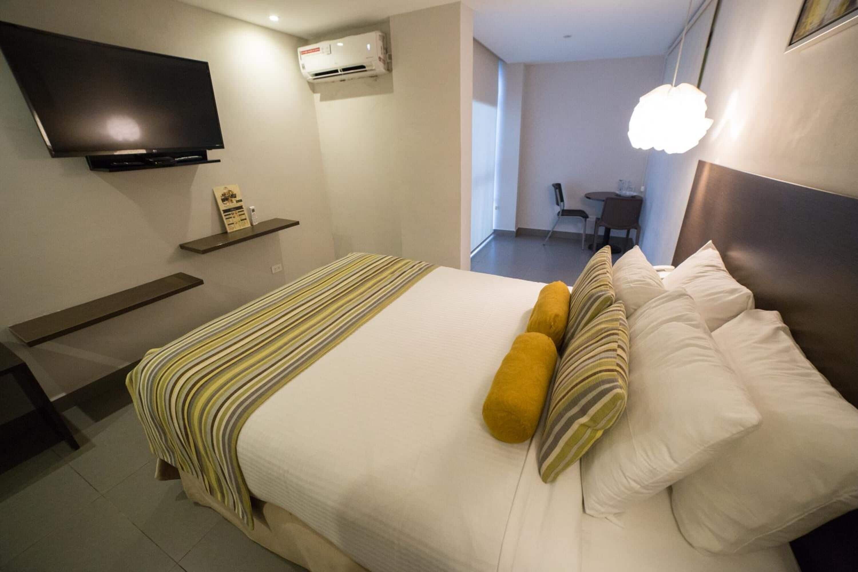 Quarto de hotel com cama, televisão na parede, mesa de cabeceira e pequena escrivaninha com cadeira.