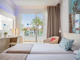 Quarto de hotel com cama, escrivaninha, cadeira e varanda com vista para palmeiras e edifícios.
