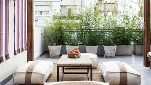 Uma área de varanda coberta com pérgola de madeira, assentos com almofadas, uma mesa com uma cesta de frutas e plantas em vasos.