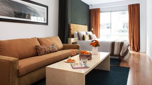 Quarto de hotel moderno com sofá, cama, mesa de centro com um livro e frutas, e uma janela grande com cortinas.