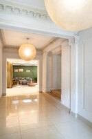 Um corredor amplo com uma luminária redonda grande, uma parede verde com padrão floral, e um espaço de estar com sofá e mesa de centro.