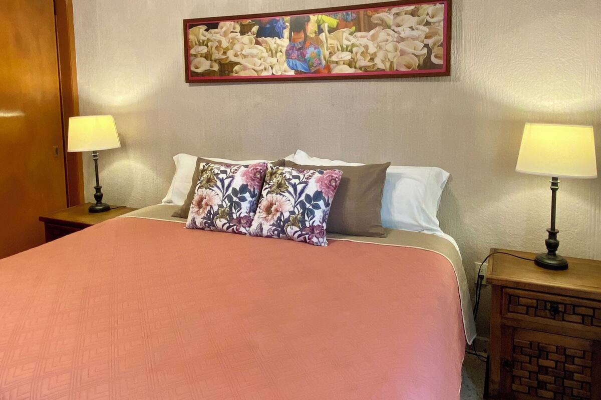 Quarto de hotel com uma cama grande, duas mesas de cabeceira com abajures, uma porta de madeira e um quadro pendurado na parede.