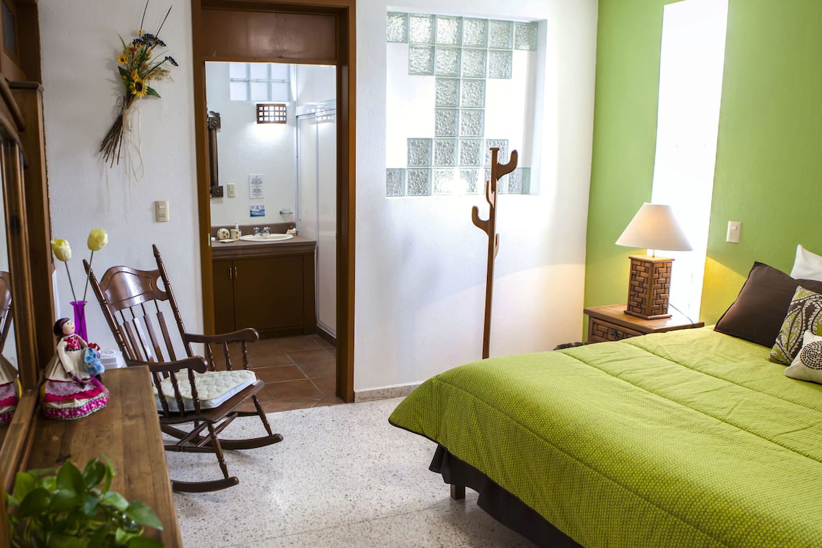 Quarto com parede verde, uma cadeira de balanço de madeira, cama com colcha verde, mesa de cabeceira com abajur e janela com persiana em forma de cruz.
