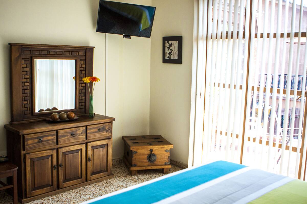 Quarto com penteadeira de madeira, televisão na parede e janela com persianas.