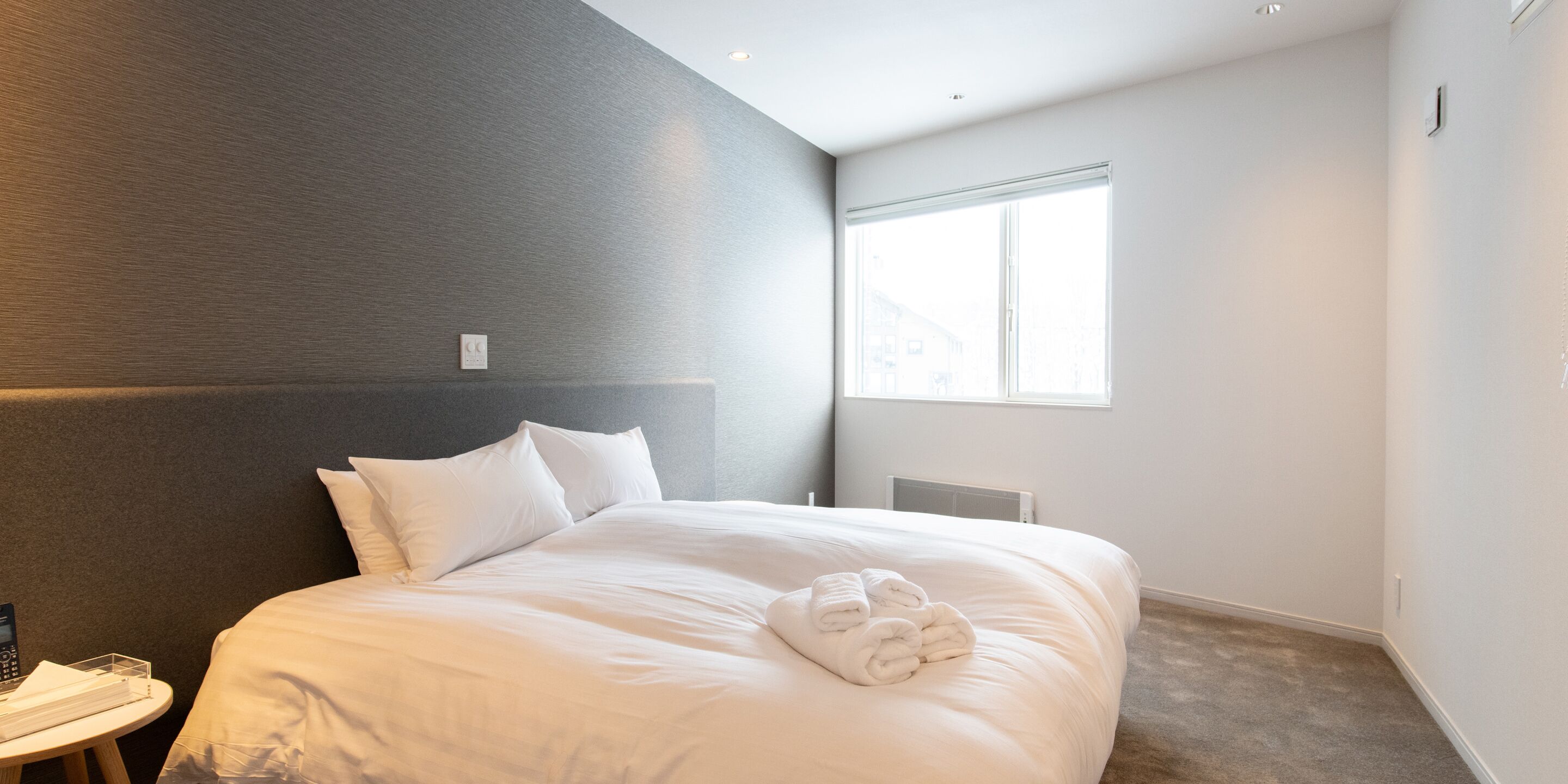 Quarto de hotel moderno com uma cama grande, mesa de cabeceira com telefone e papéis, e uma janela com vista para prédios.