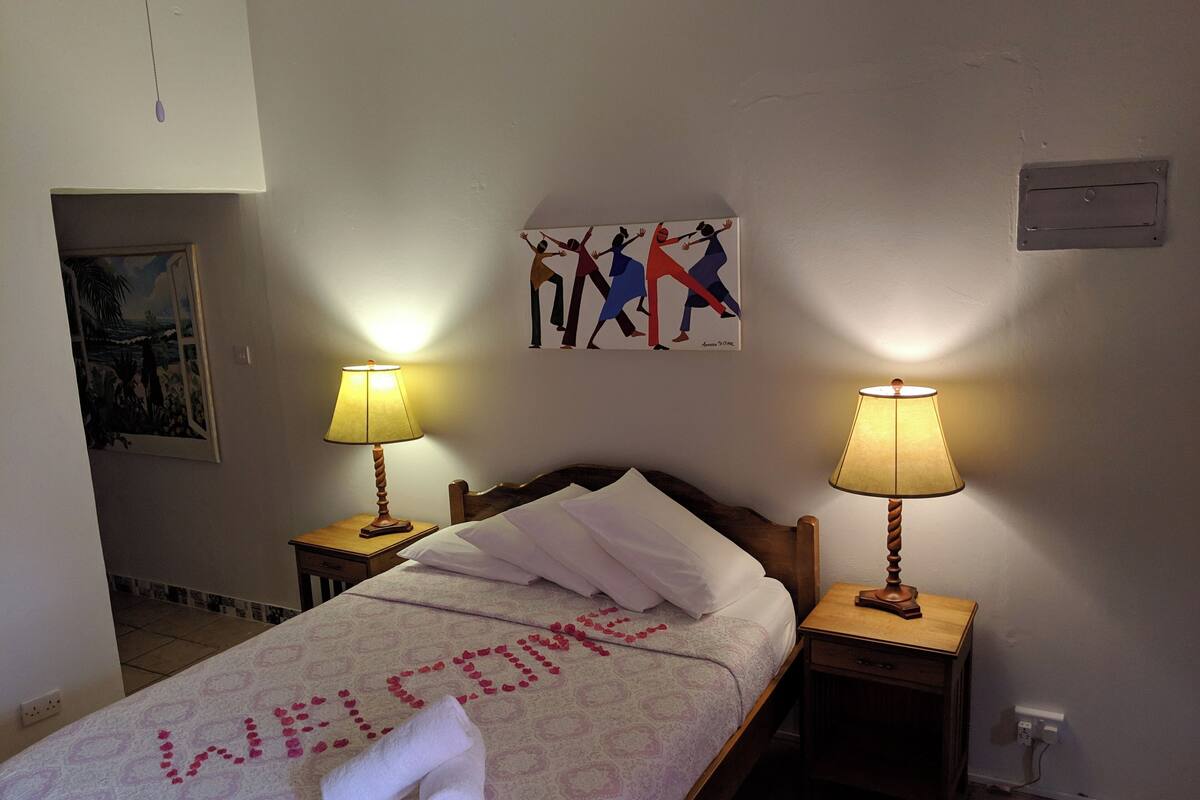 Cama bem arrumada, tapete de boas-vindas, duas luminárias de mesa, um quadro na parede e uma tomada.