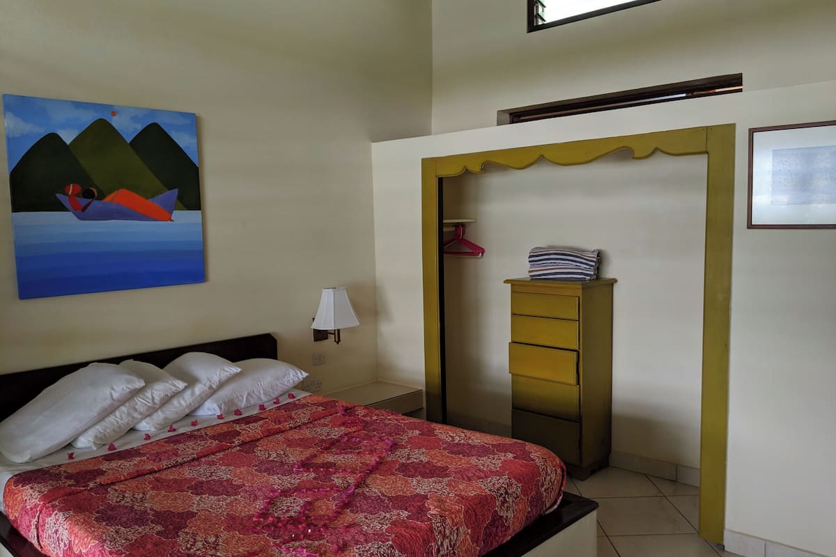Quarto com cama, quadro, penteadeira e porta que dá para outro cômodo.
