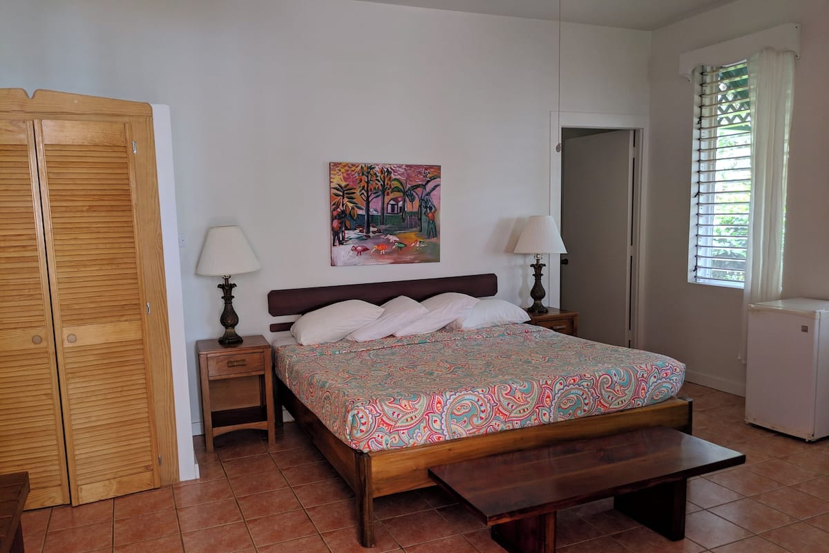 Quarto com cama de madeira, duas mesas de cabeceira com abajures, um banco, um quadro colorido na parede e uma janela com persianas.