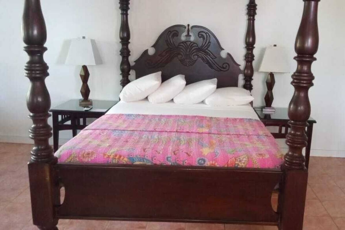 Uma cama de dossel com colcha florida, duas mesas de cabeceira com abajures, e uma parede lisa ao fundo.