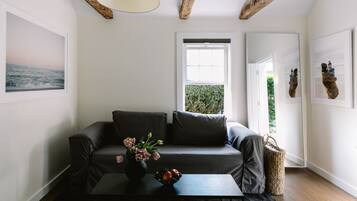 Uma sala de estar moderna com um sofá de couro escuro, uma mesa de centro preta com um vaso e frutas, uma janela grande com vista para o verde e um vigamento de madeira no teto.