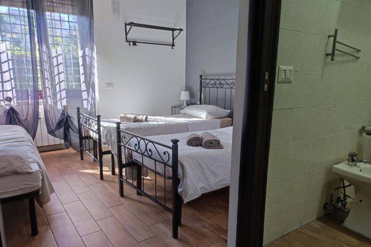Quarto com duas camas, mesa de cabeceira, prateleira suspensa e banheiro com pia e vaso sanitário.