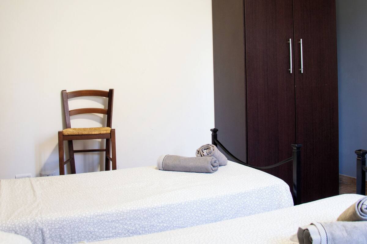 Quarto com duas camas, uma cadeira de madeira e um guarda-roupa de madeira escura.