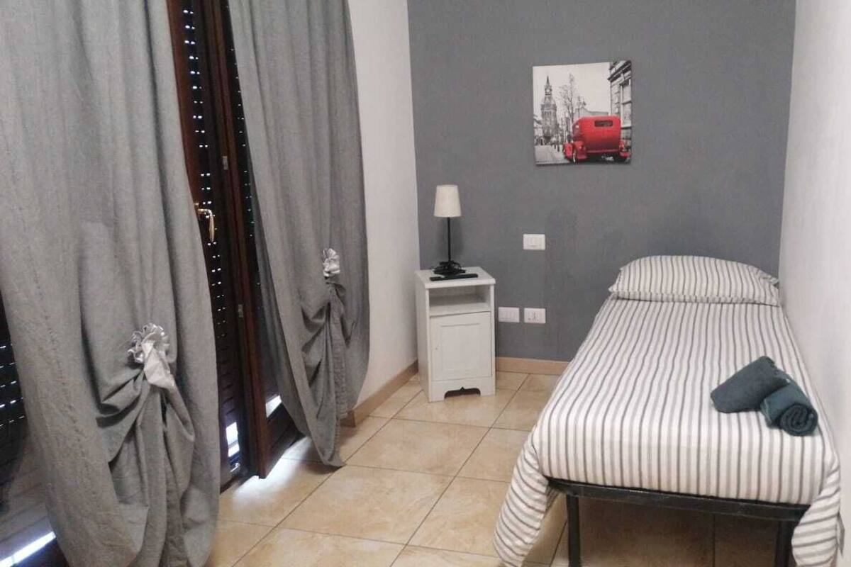 Quarto com cama, mesa de cabeceira, luminária, janela com cortinas e um quadro na parede.