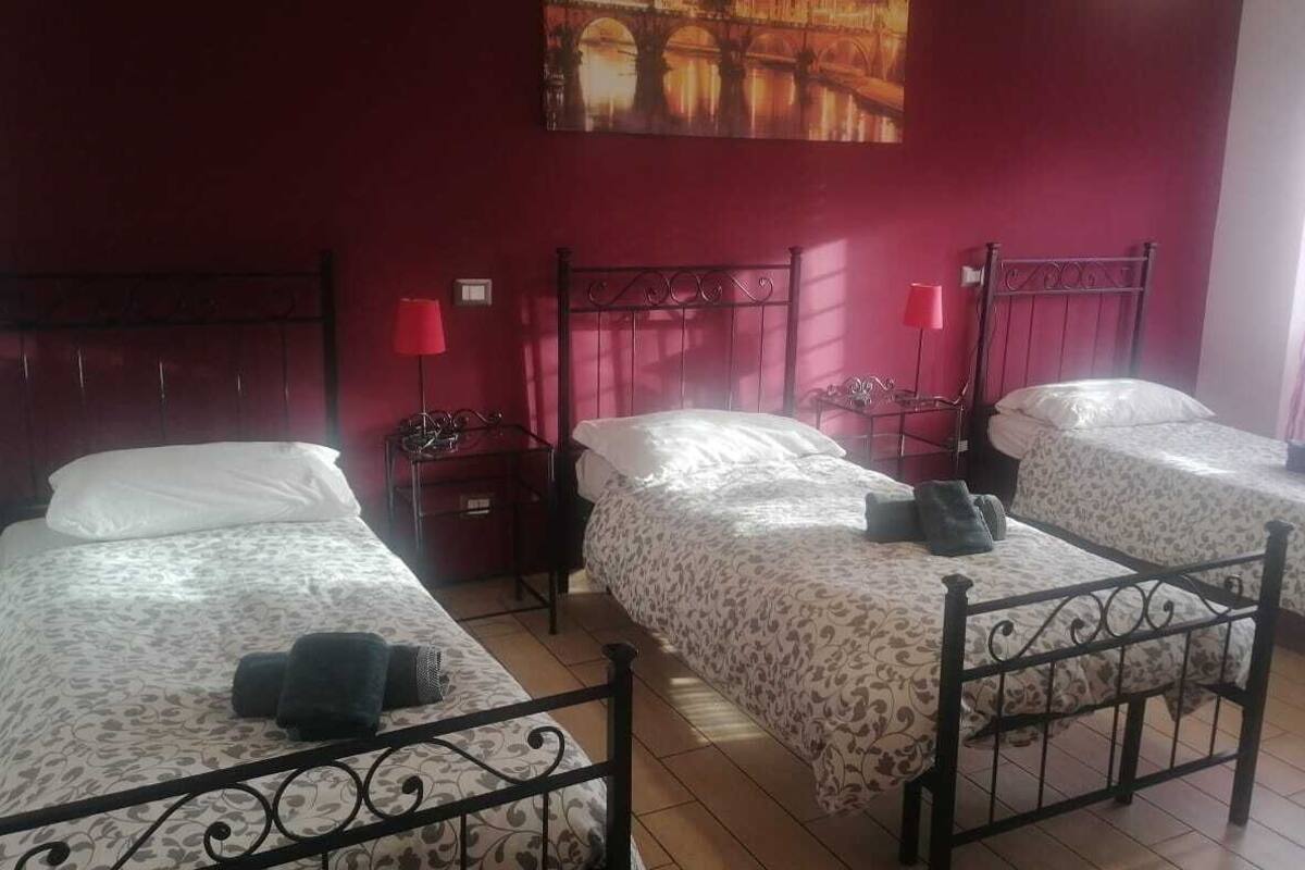 Quarto de hotel com três camas, cada uma com estrutura de metal e roupa de cama estampada. Há mesas de cabeceira com luminárias e uma parede vermelha com um quadro.