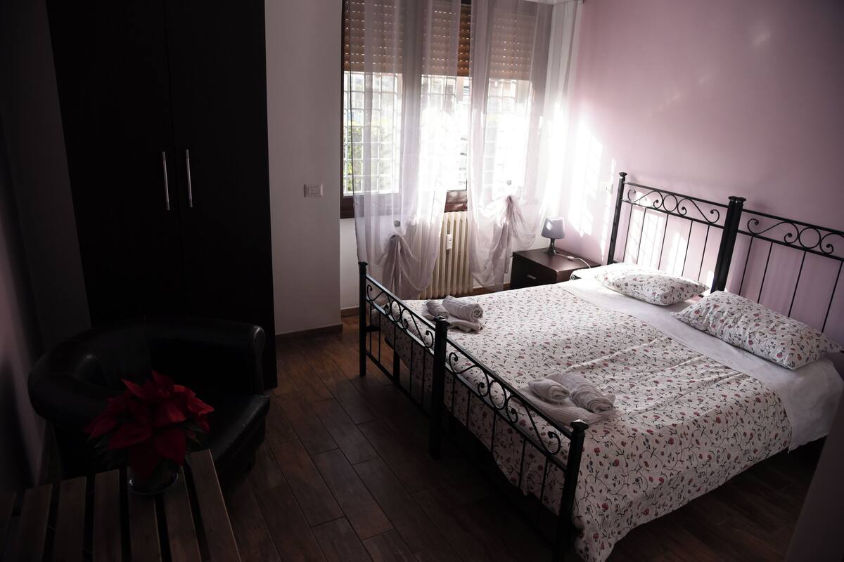 Quarto com cama, cadeira, mesa de cabeceira e janela com cortinas.
