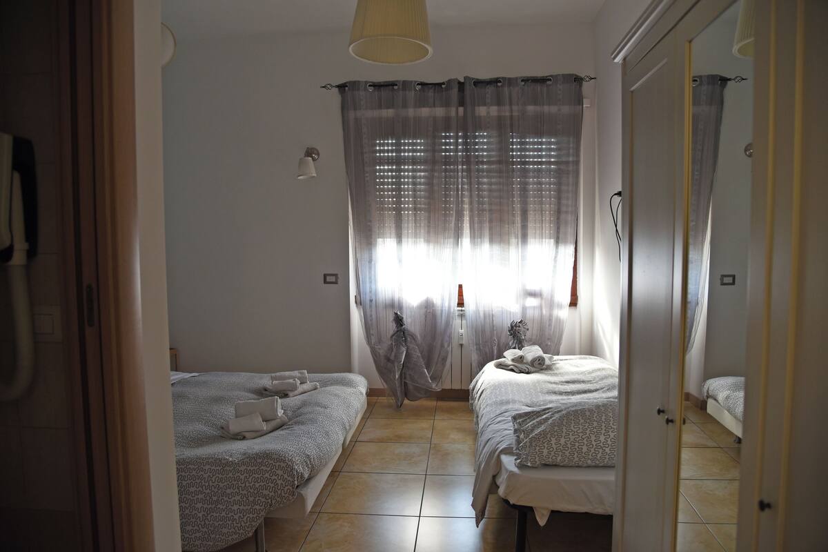 Quarto com duas camas, uma janela com cortinas leves e um espelho.
