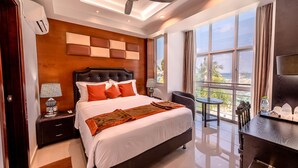 Quarto de hotel moderno com uma cama grande, um ventilador de teto, uma mesa de cabeceira com abajur, uma escrivaninha com cadeira e vista para o oceano através de janelas amplas.