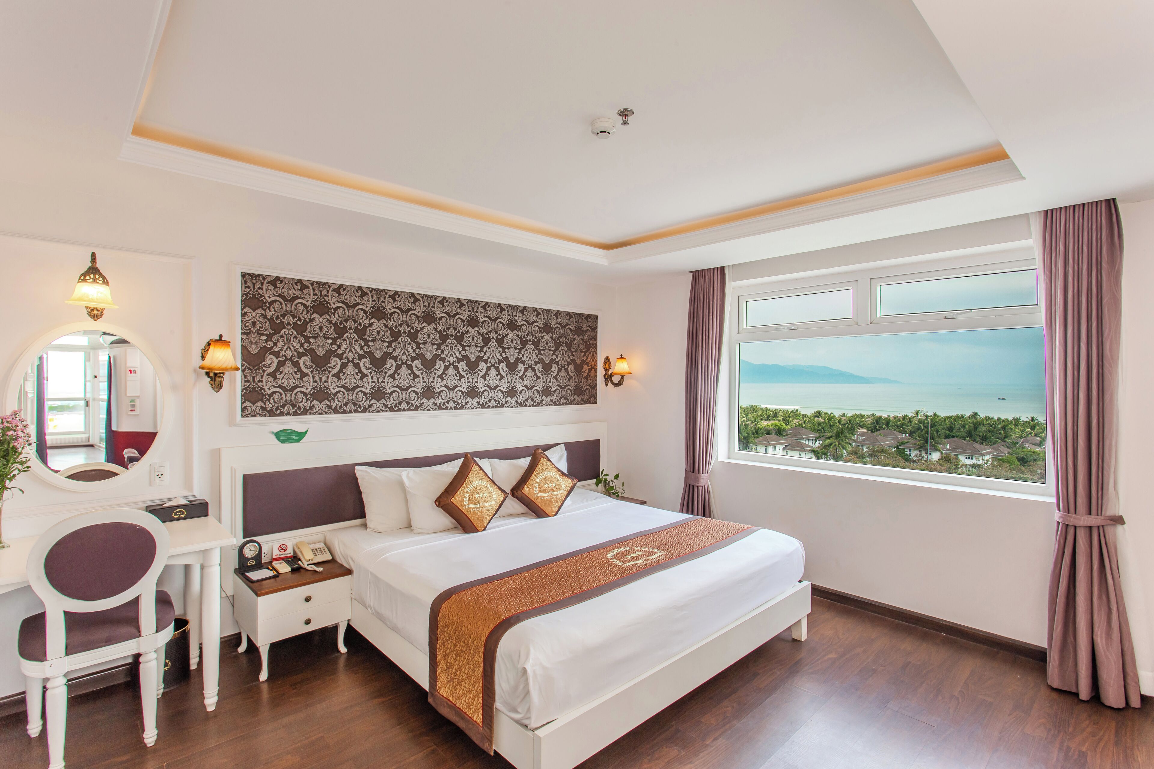 Quarto de hotel com uma cama grande, uma escrivaninha com cadeira, uma toucador com espelho e vista para o mar.