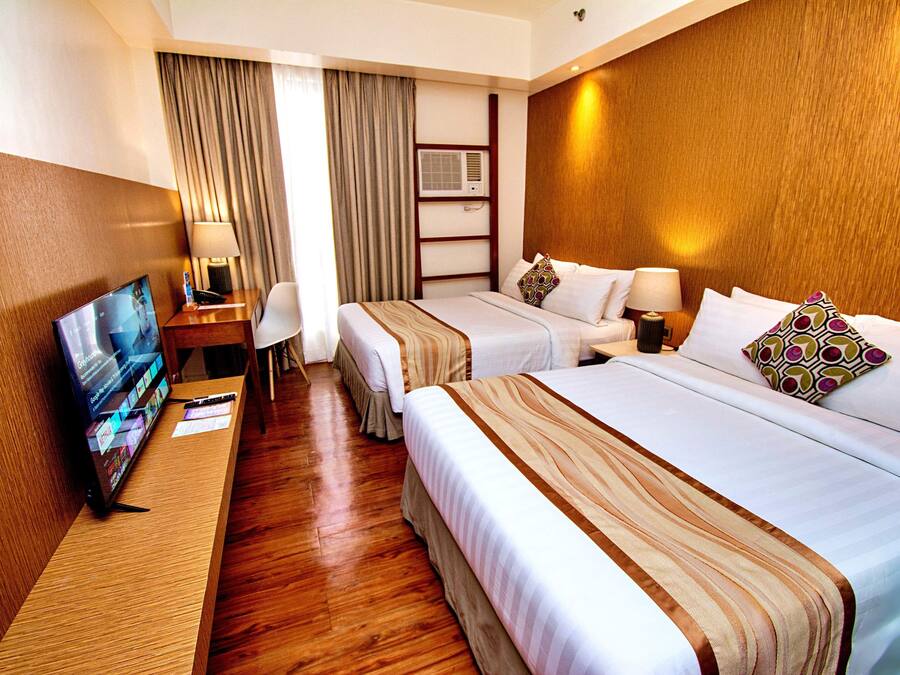 Quarto de hotel com piso de madeira, uma TV sobre suporte de madeira, duas camas com lençóis brancos e cobertores listrados, uma escrivaninha com luminária e uma janela com cortinas.