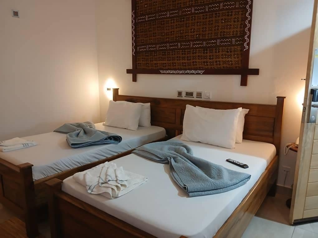 Quarto de hotel com duas camas de solteiro, cabeceira de madeira, luminária fixada na parede e tapeçaria tecida sobre as camas.