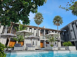 Área da piscina com espreguiçadeiras e guarda-sóis, uma piscina e um edifício com varandas.