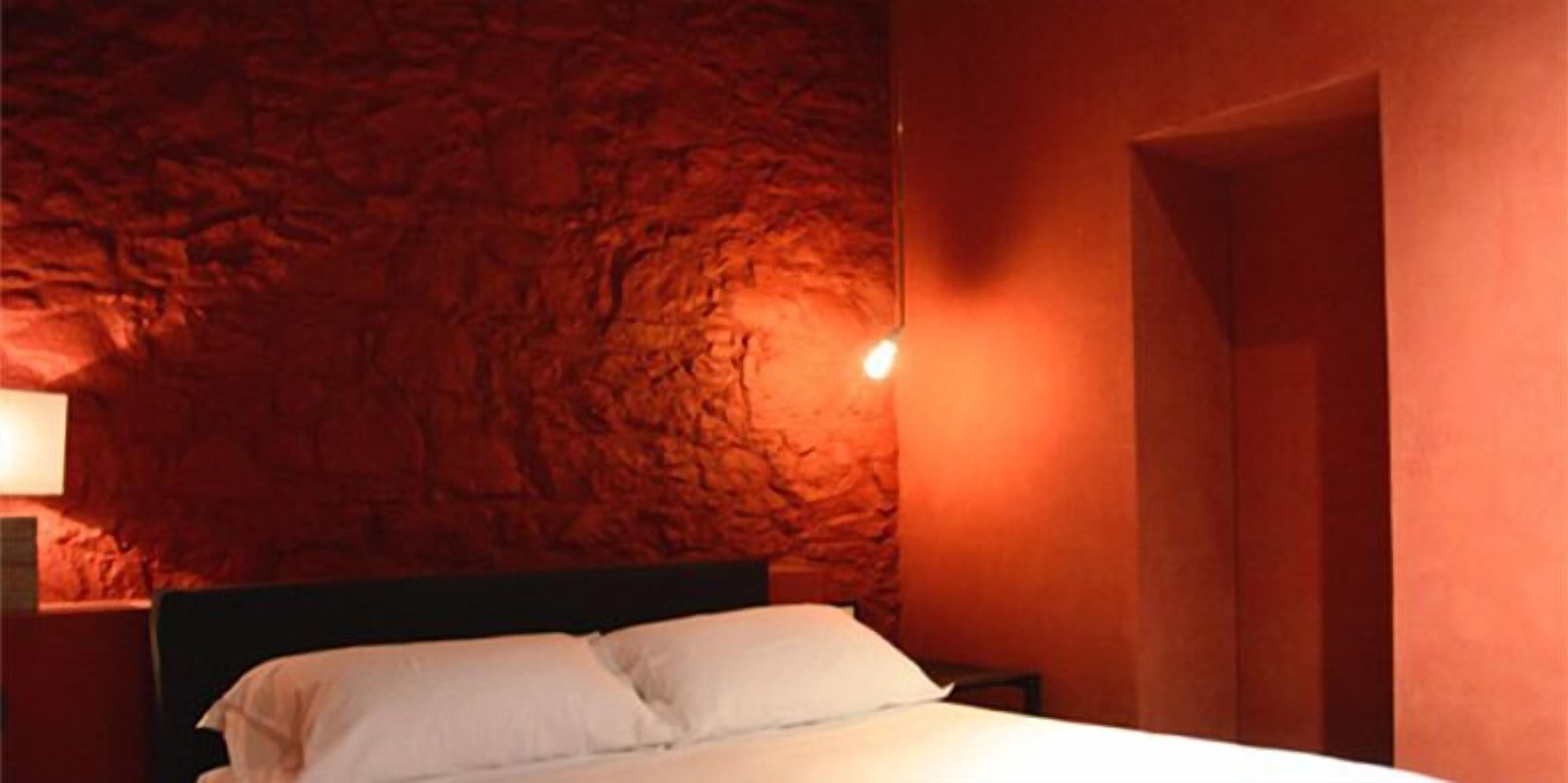 Quarto com parede de pedra, cama com roupa de cama branca e uma luminária de cabeceira.