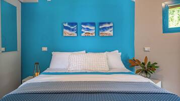 Um quarto com parede azul, cama com roupa de cama branca e estampada, três quadros pendurados na parede, mesa de cabeceira com luminária e uma planta em vaso.
