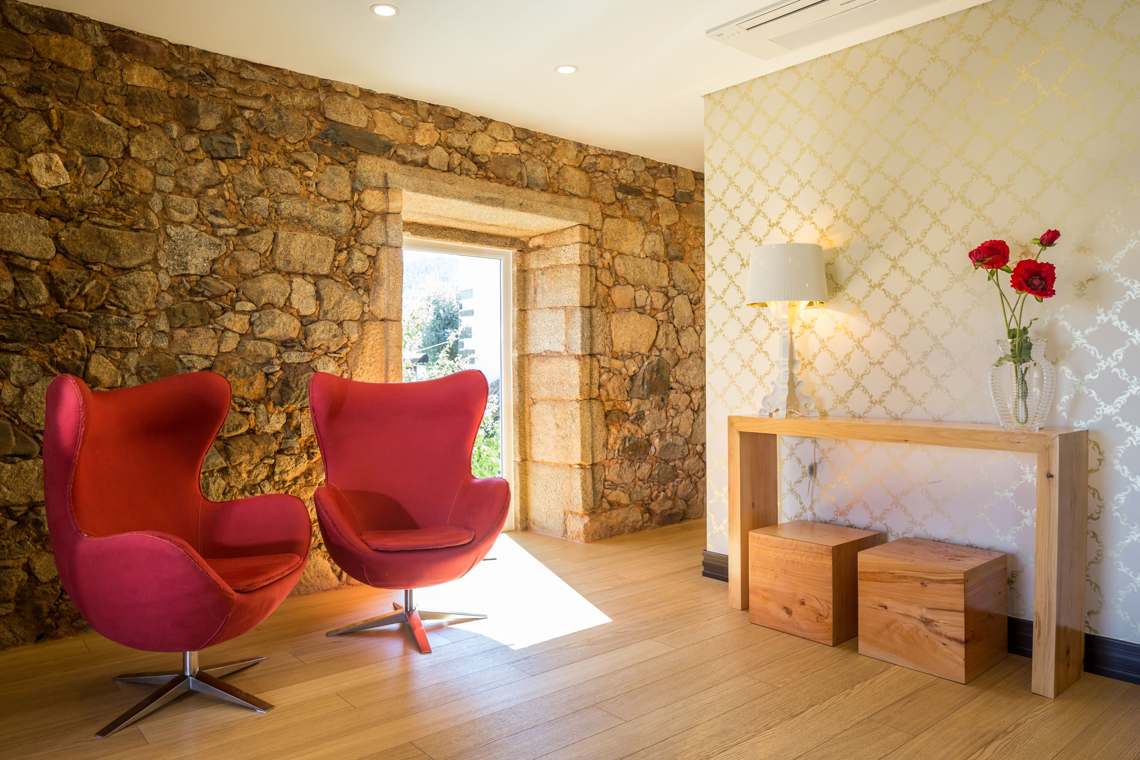 Quarto com parede de pedra, duas cadeiras vermelhas, mesa consola de madeira com abajur e um vaso com flores.