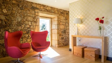 Quarto com parede de pedra, duas cadeiras vermelhas, mesa consola de madeira com abajur e um vaso com flores.