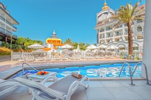 Área da piscina com espreguiçadeiras, uma mesa com frutas, e vista para um hotel com uma cúpula amarela característica e varandas brancas.