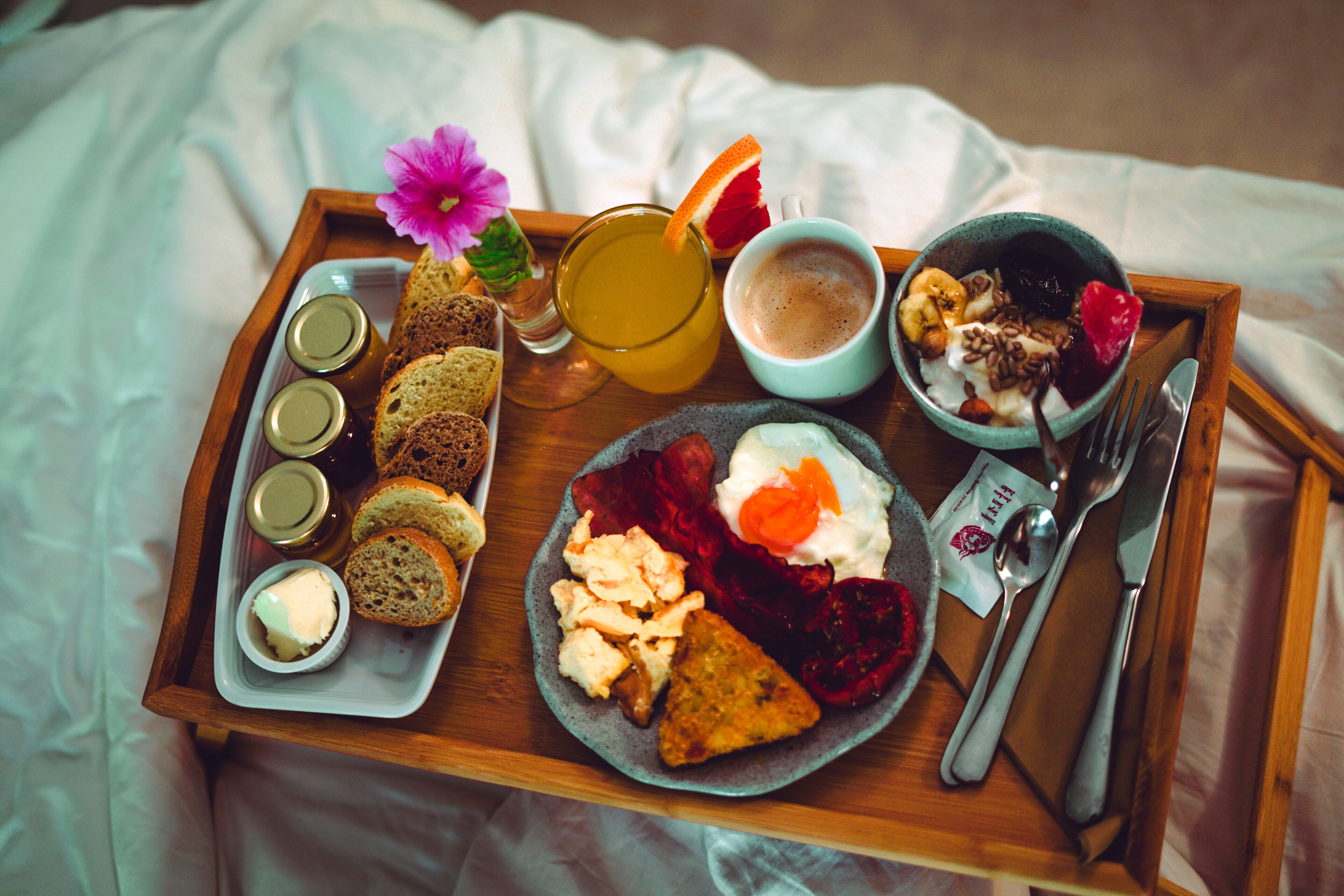 Uma bandeja de café da manhã com torradas, geleia, manteiga, uma xícara de café, uma tigela de iogurte com frutas e um copo de suco sobre uma cama.