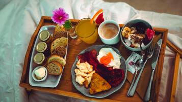 Uma bandeja de café da manhã com torradas, geleia, manteiga, uma xícara de café, uma tigela de iogurte com frutas e um copo de suco sobre uma cama.