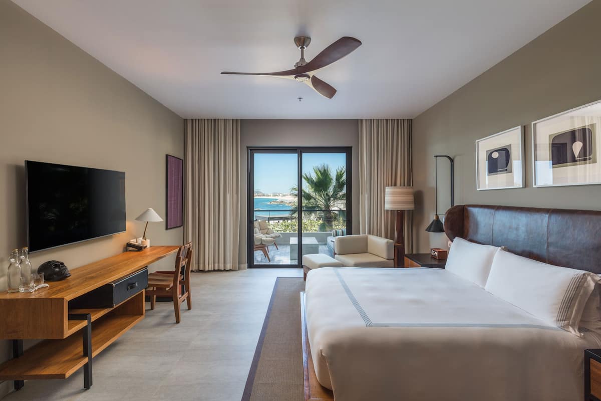 Quarto de hotel com cama, escrivaninha, televisão, ventilador de teto, janela com vista e porta para uma varanda.