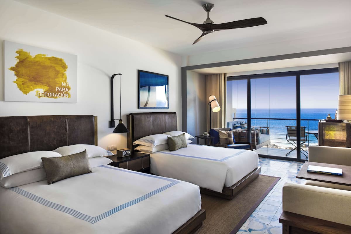 Quarto de hotel com duas camas, um ventilador de teto, uma janela grande com vista para o oceano e obras de arte nas paredes.