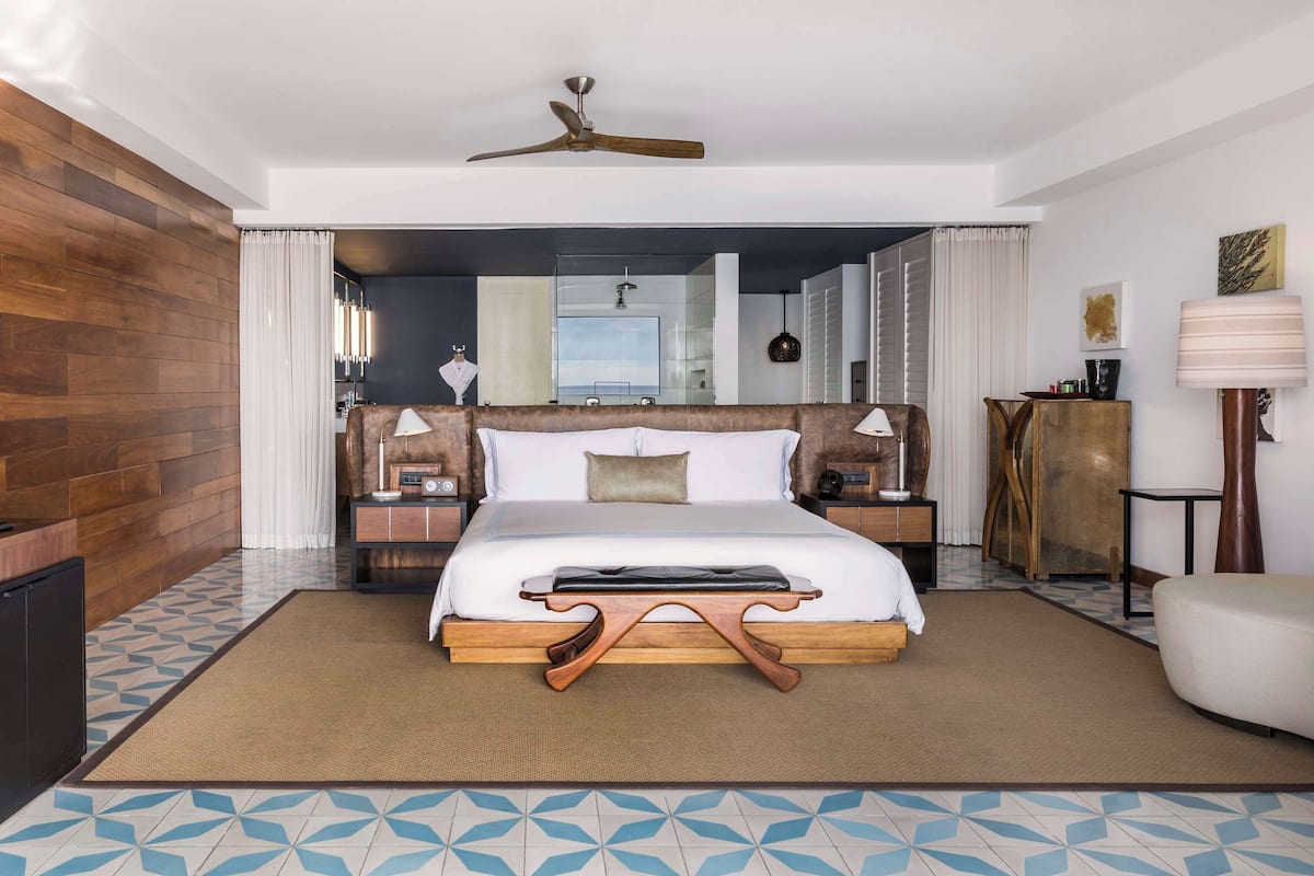 Quarto de hotel moderno com uma cama grande, cabeceira de madeira, mesas de cabeceira com luminárias, um guarda-roupa e um ventilador de teto.