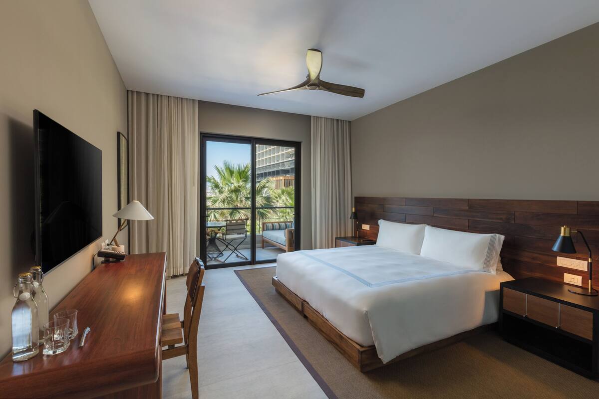 Quarto de hotel com uma cama grande, televisão de tela plana, escrivaninha com abajur e varanda com vista para palmeiras.