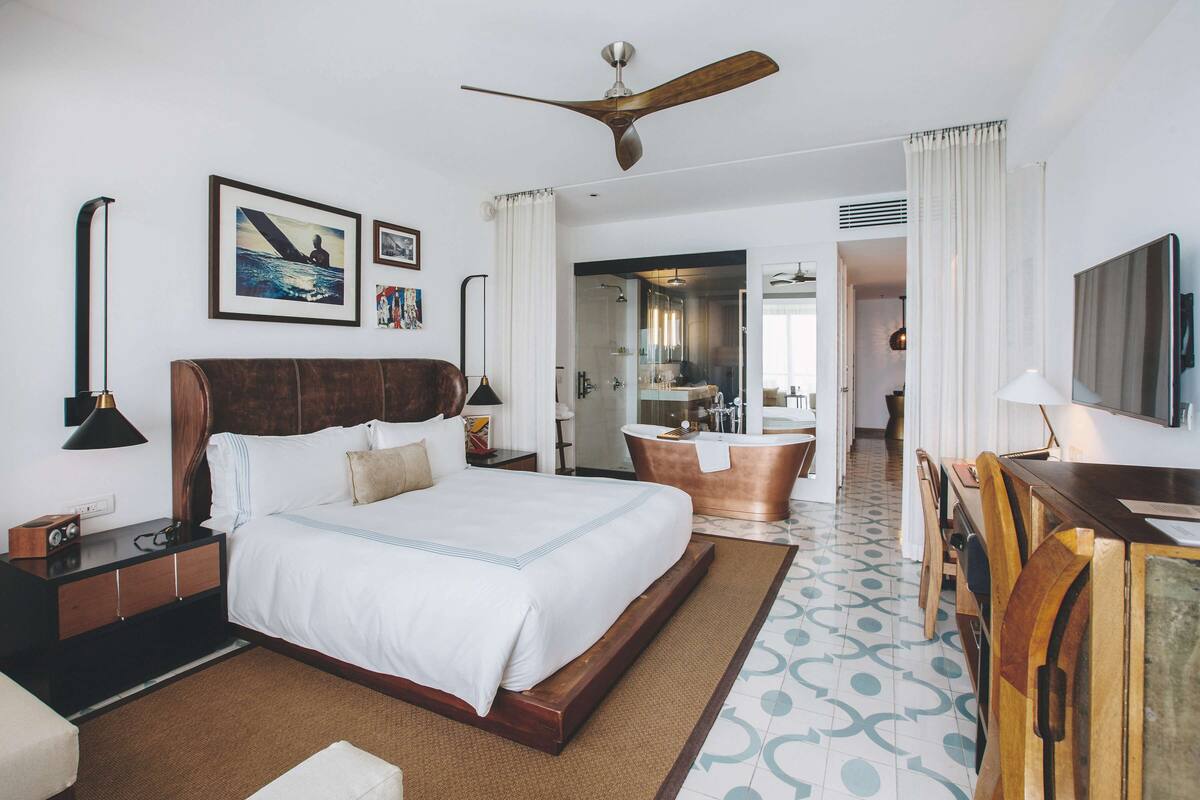 Quarto de hotel com uma cama grande, cabeceira de madeira, ventilador de teto, banheiro com banheira e área de jantar com mesa e cadeiras.