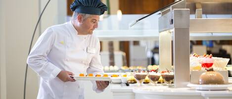 Cozinheiro com uniforme branco segurando uma bandeja de sobremesas em um buffet.