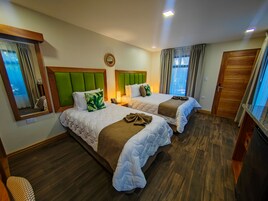 Quarto de hotel com duas camas, cabeceiras de madeira, um espelho, mesa de cabeceira com abajur e vista para o exterior pela janela.