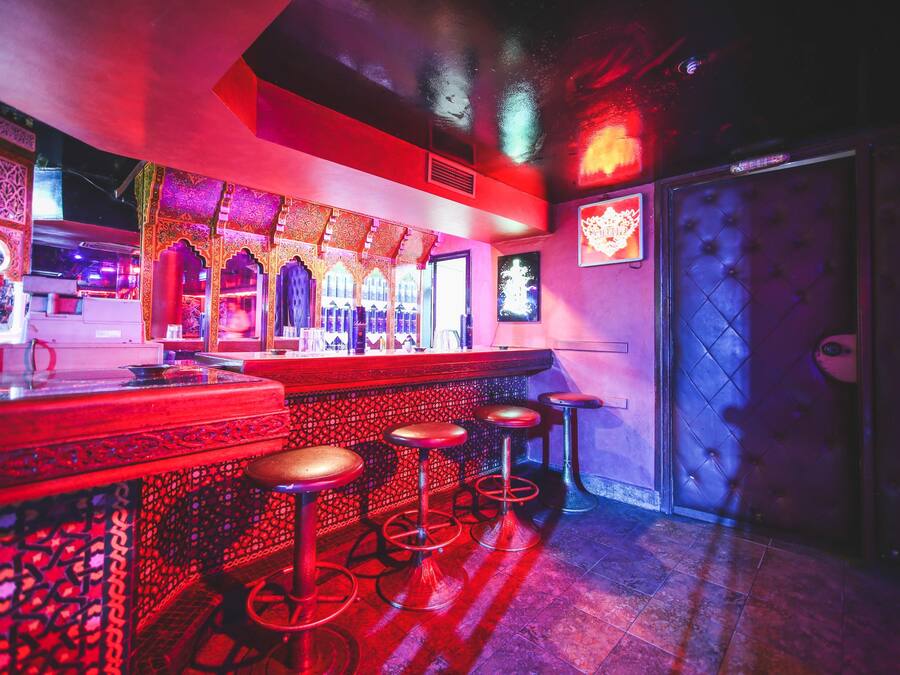 Uma área de bar com iluminação ornamentada em vermelho e roxo, um balcão e vários bancos.