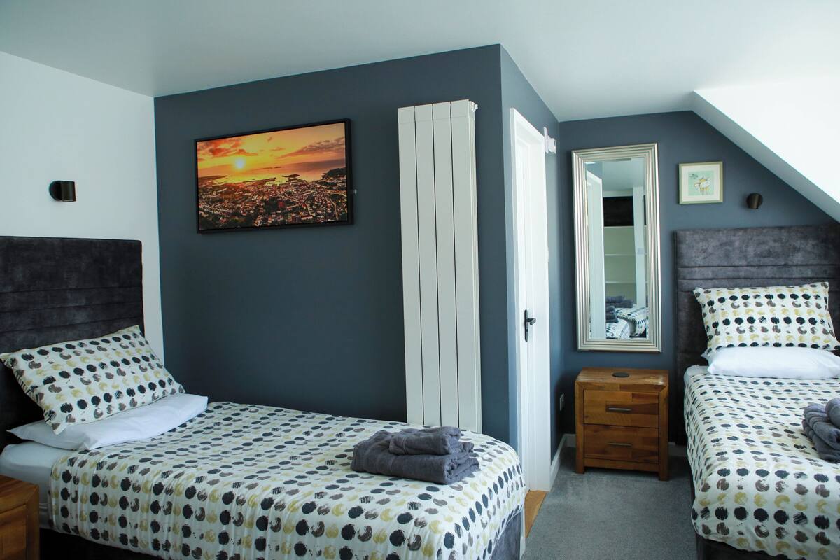 Quarto com duas camas, uma pintura de paisagem urbana, um radiador branco, um espelho e uma mesinha de cabeceira de madeira.