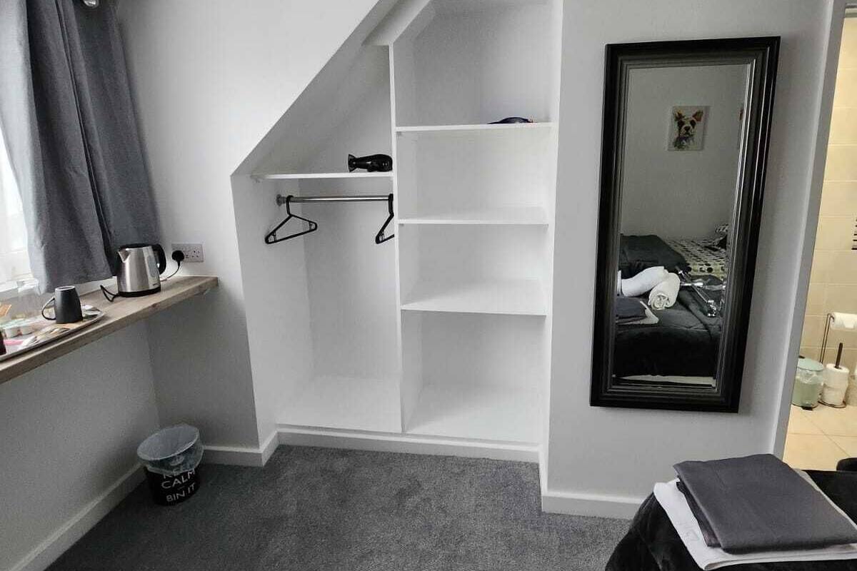 Quarto compacto com roupeiro embutido, secretária pequena com chaleira e cama com cobertura escura.