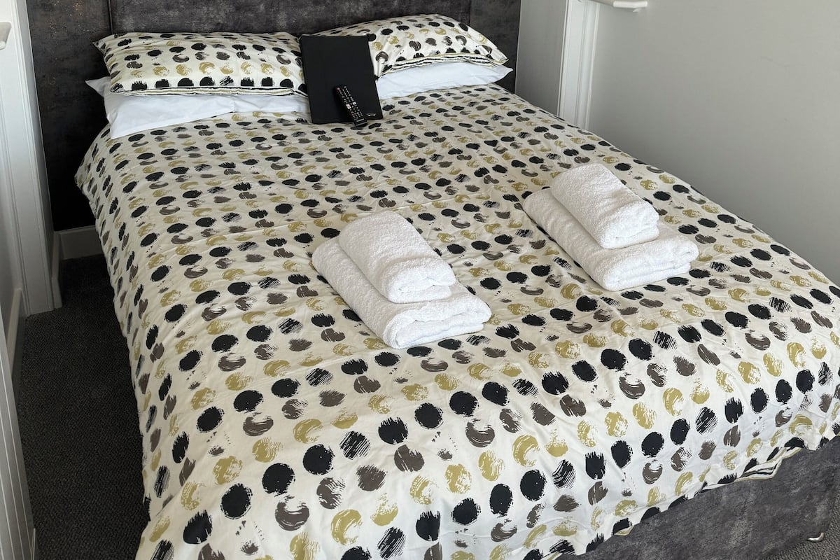 Cama bem arrumada com colcha de bolinhas, duas almofadas e duas toalhas dobradas.
