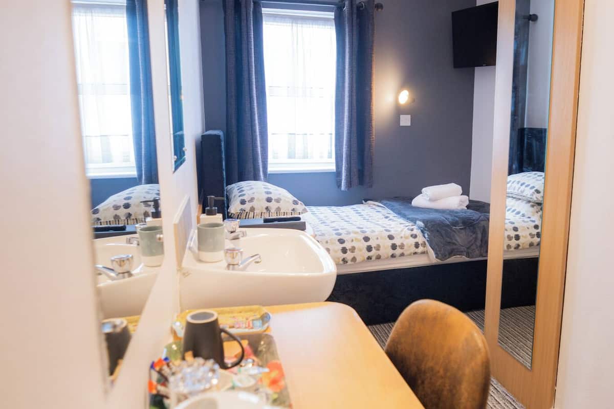 Quarto de hotel com cama, escrivaninha com chaleira, espelho, TV fixada na parede e janela com cortinas.