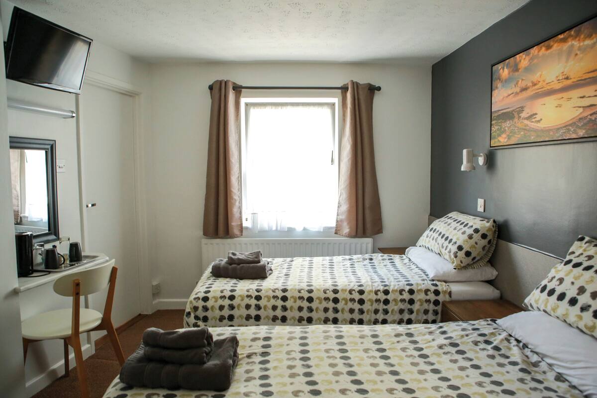 Quarto de hotel com duas camas, uma escrivaninha, uma cadeira, uma janela com cortinas e um quadro na parede.