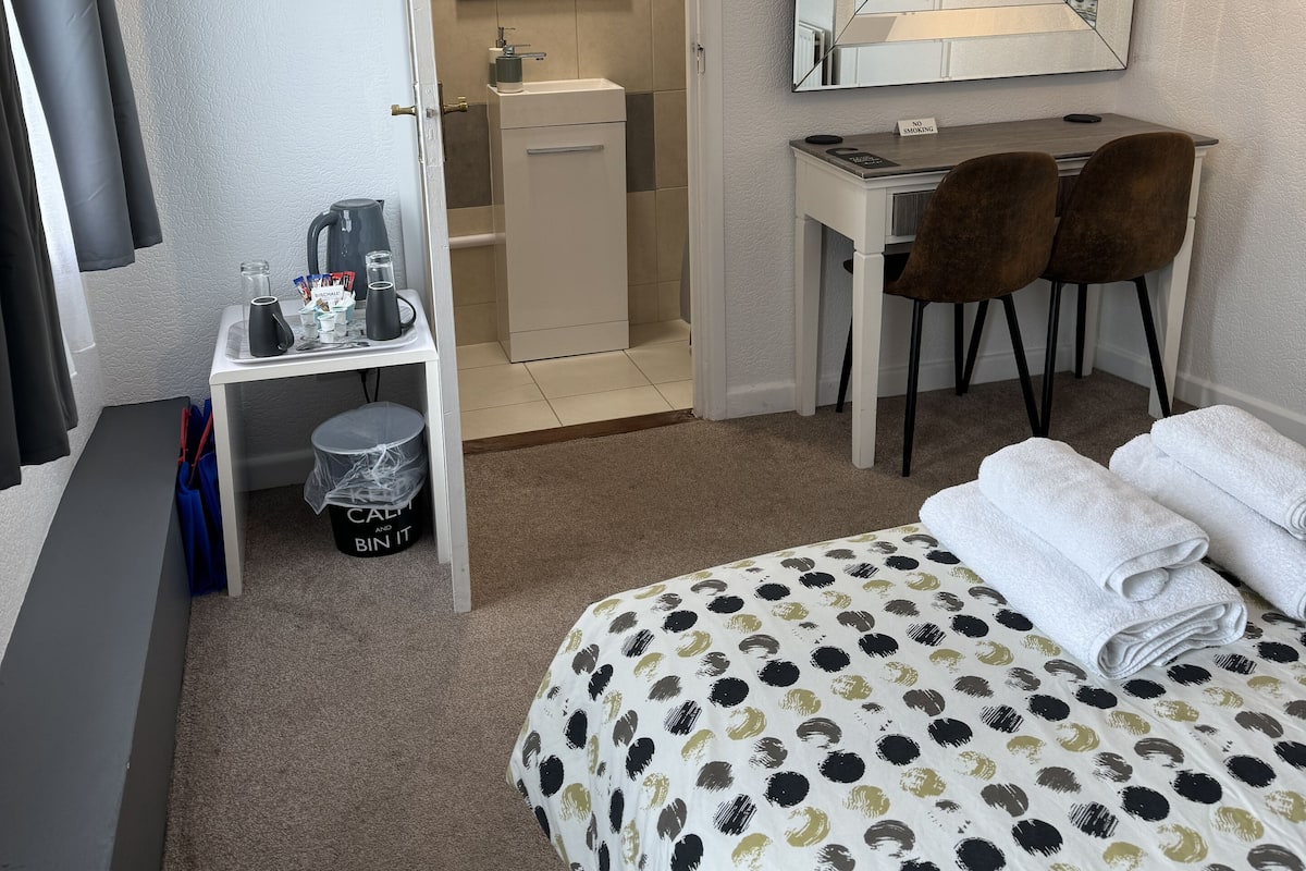 Quarto de hotel com cama, escrivaninha com duas cadeiras, televisão, espelho e banheiro visível por uma porta aberta.