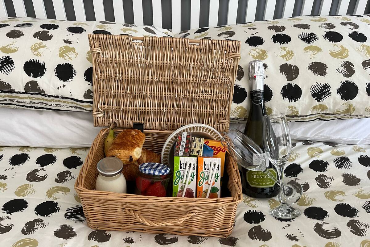 Uma cesta de vime com alimentos e uma garrafa de vinho sobre uma cama com almofadas estampadas.