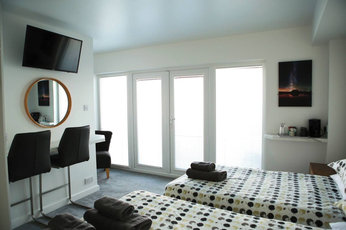 Quarto de hotel com televisão, escrivaninha, duas camas com roupa de cama em bolinhas, espelho redondo e uma janela grande.