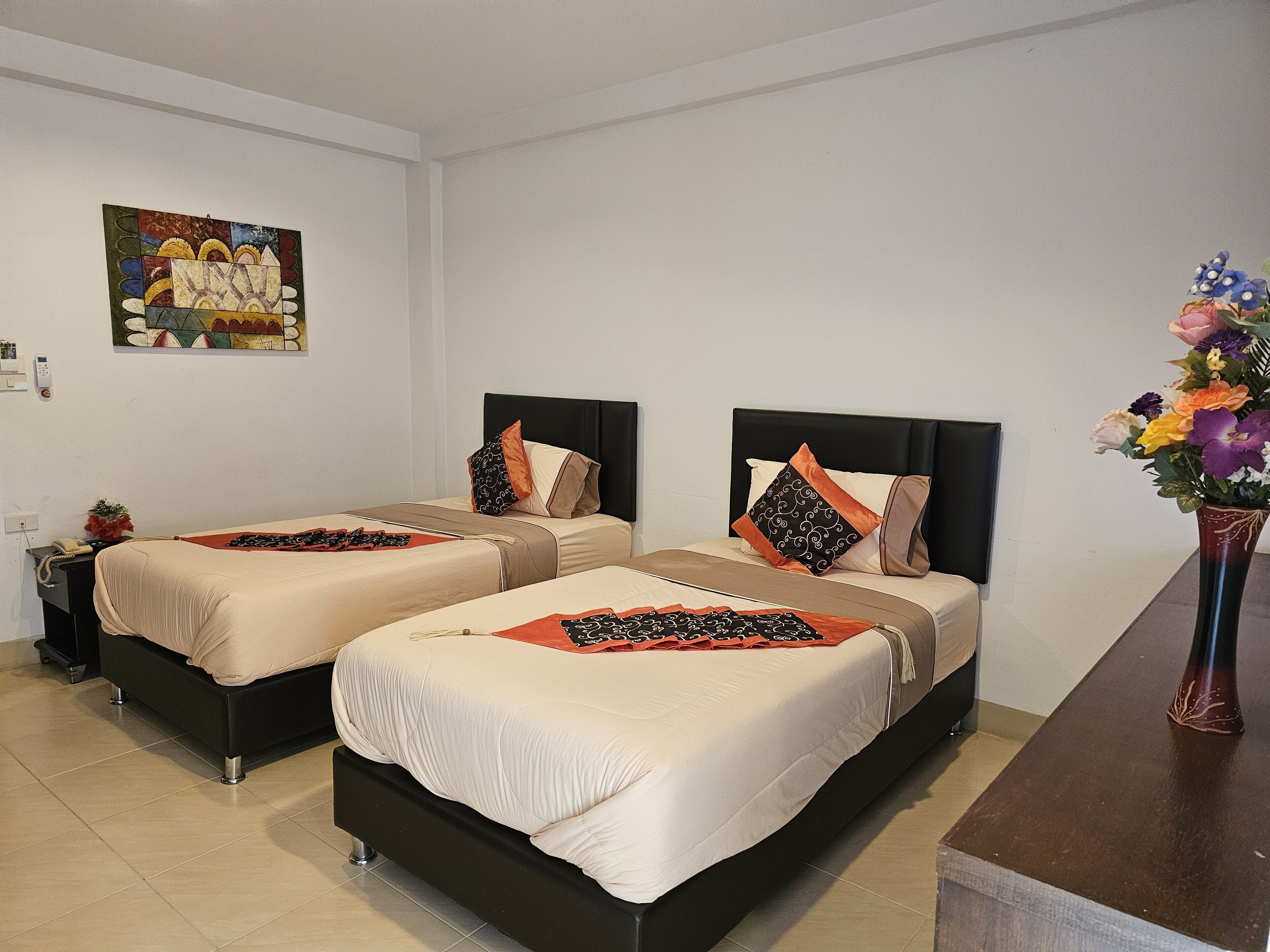 Quarto de hotel com duas camas, cada uma com cabeceira preta, roupa de cama bege e almofadas decorativas. Há uma pequena mesa com um vaso de flores e um quadro na parede.